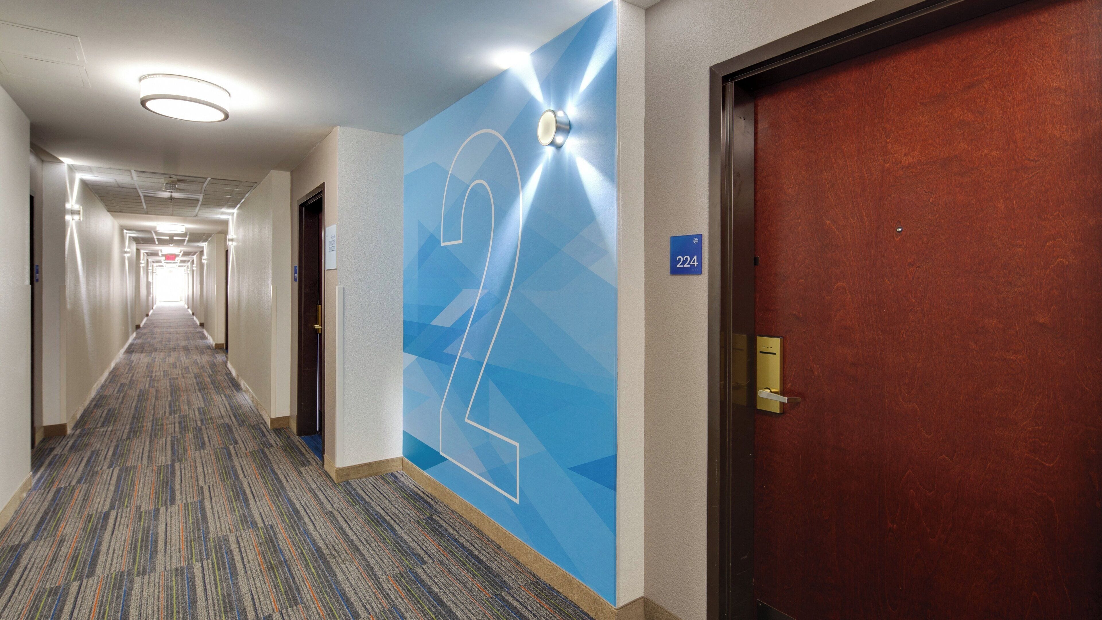 Corredor de hotel com pisos carpetados, parede azul com design geométrico e porta com placa de número.