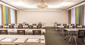 Uma sala de conferências com fileiras de mesas e cadeiras, um candelabro e cortinas estampadas.