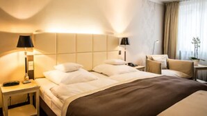 Quarto de hotel com cama, mesas de cabeceira, luminárias, uma cadeira e janela com cortinas.