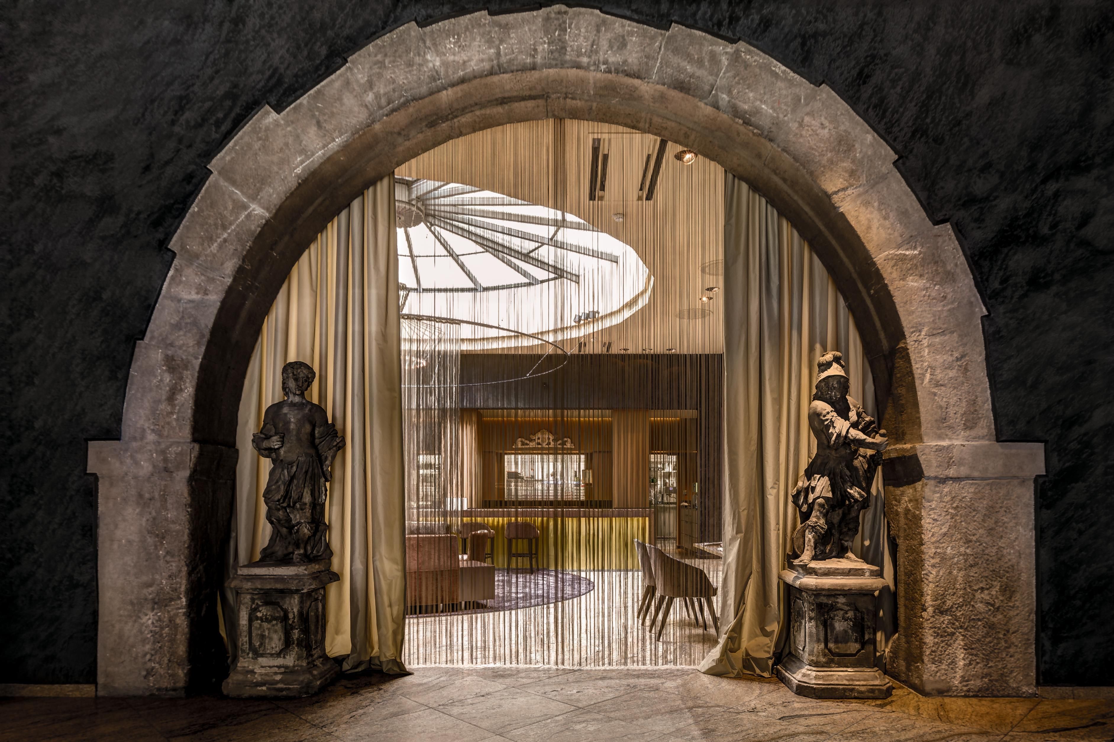 Uma porta arqueada com duas estátuas em pedestais, uma mesa circular e uma cortina estampada.