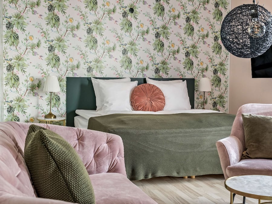 Quarto com papel de parede floral, colcha verde e mesa redonda.