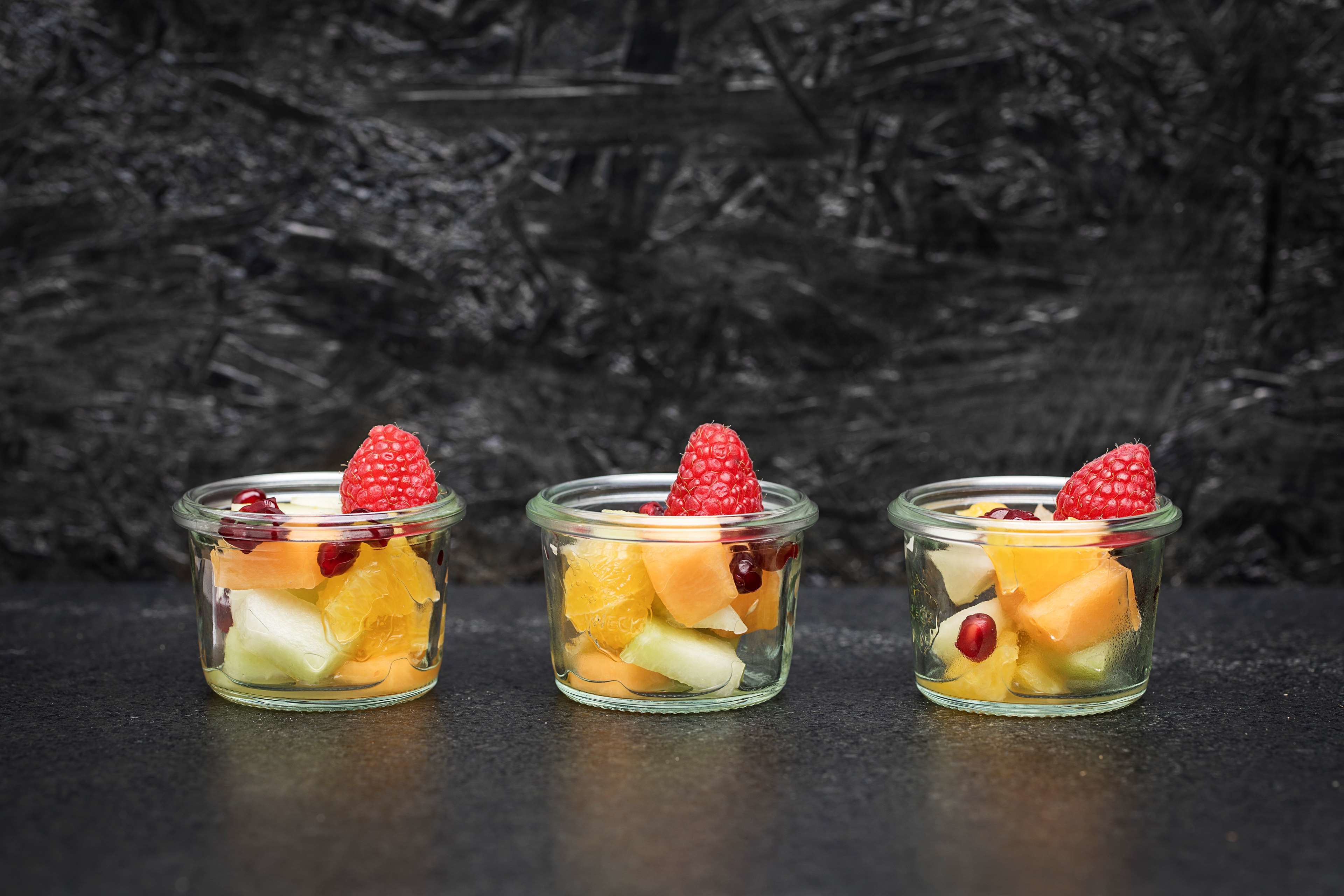 Três recipientes de vidro com salada de frutas, cada um com uma morango e um framboesa, sobre uma superfície escura.