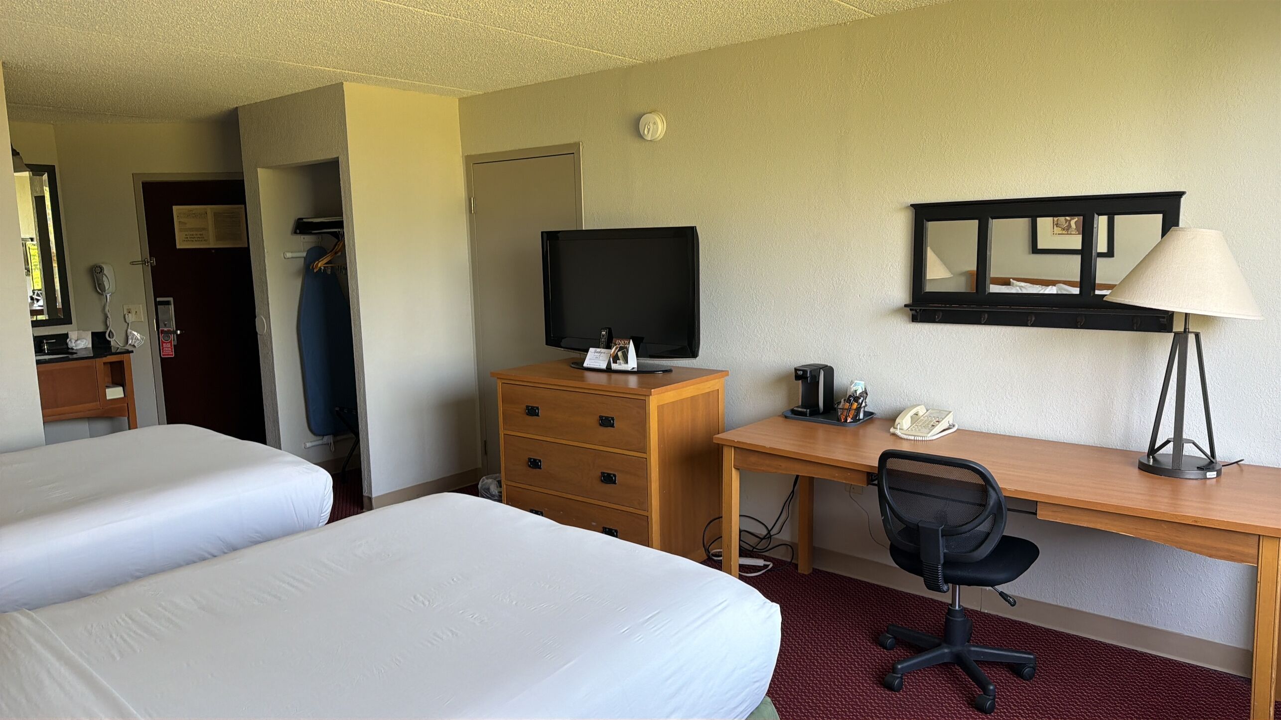 Quarto de hotel com duas camas, uma escrivaninha com cadeira, televisão, luminária e telefone.
