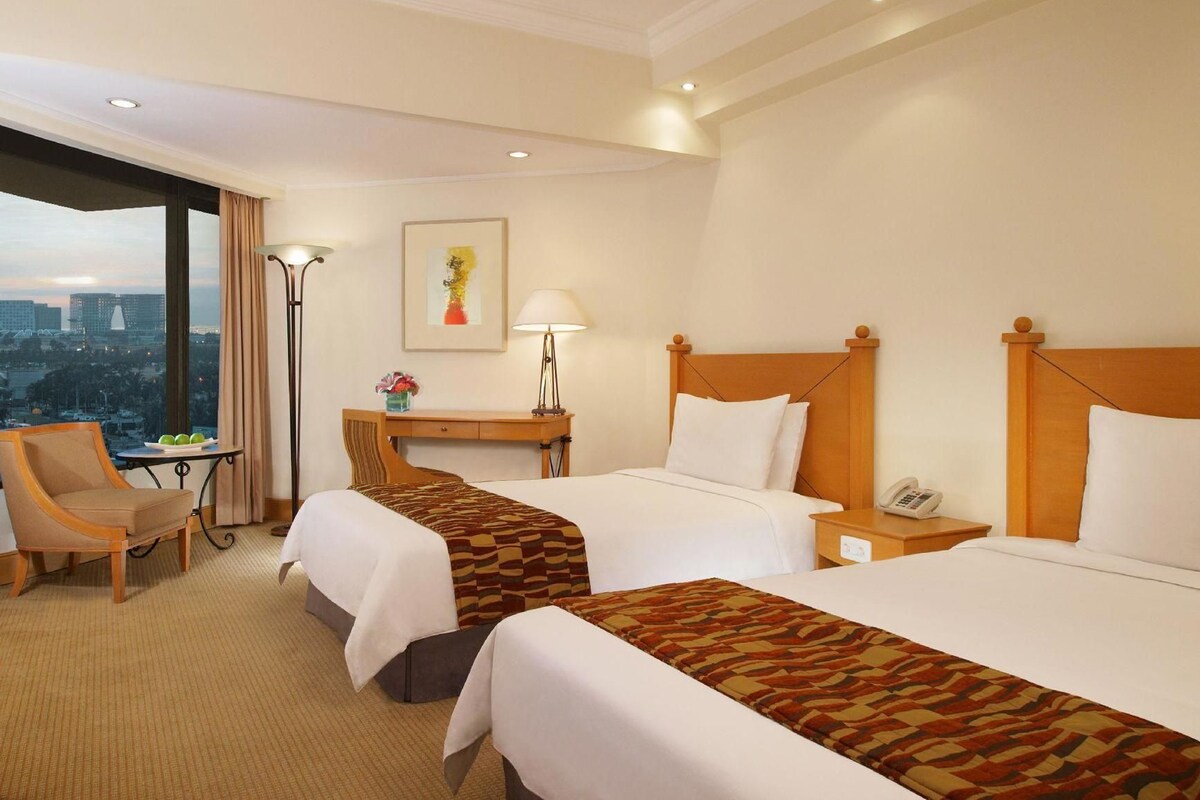 Quarto de hotel com duas camas, uma escrivaninha, uma cadeira, um abajur e vista para a cidade.
