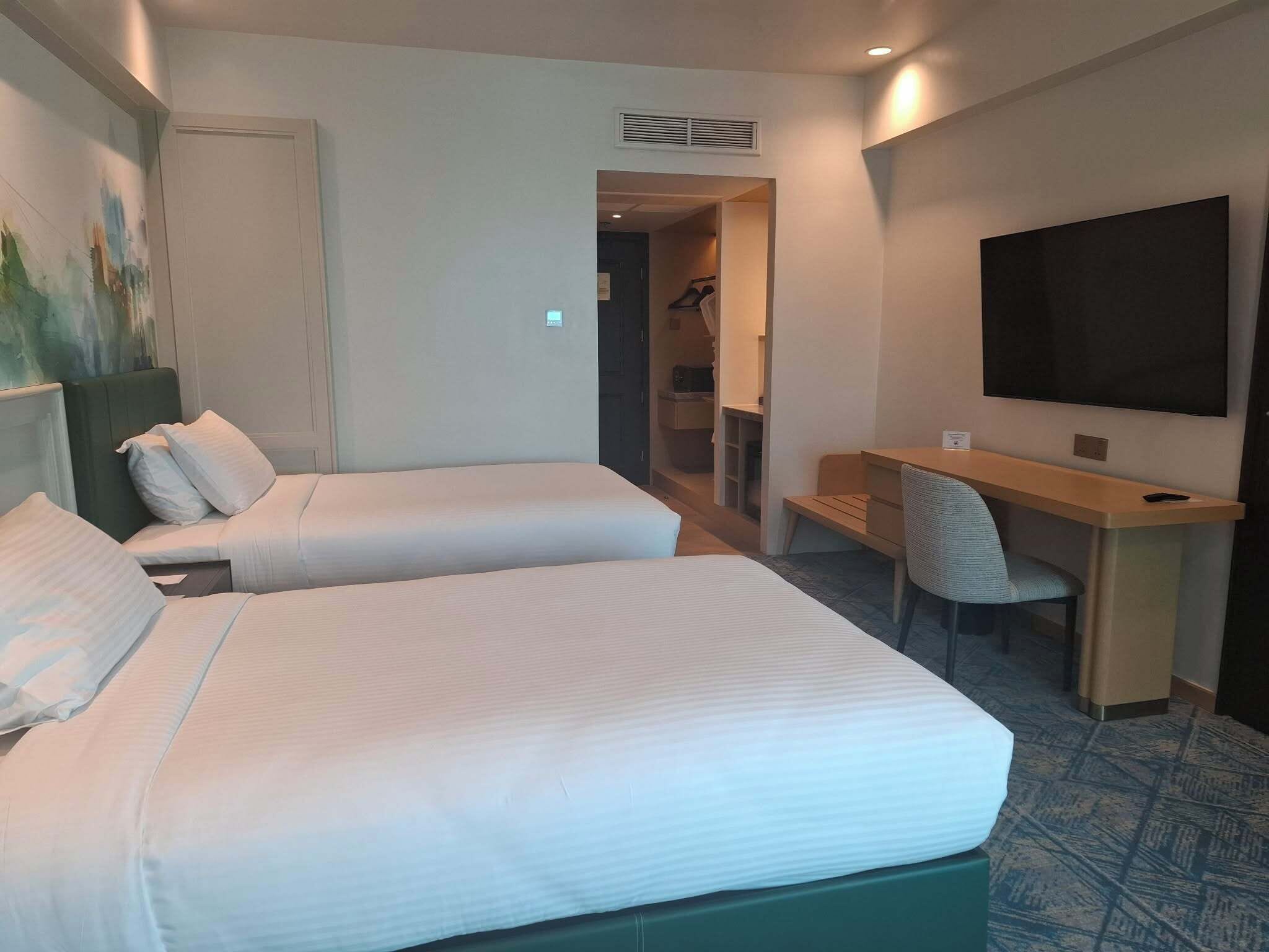 Quarto de hotel com duas camas, uma escrivaninha, uma cadeira, uma televisão e um quadro na parede.