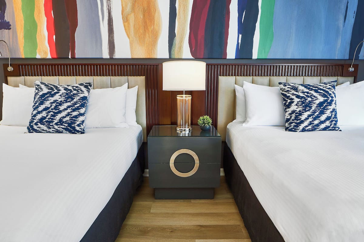 Quarto de hotel com duas camas, uma mesa de cabeceira com abajur e uma pintura abstrata colorida sobre as camas.