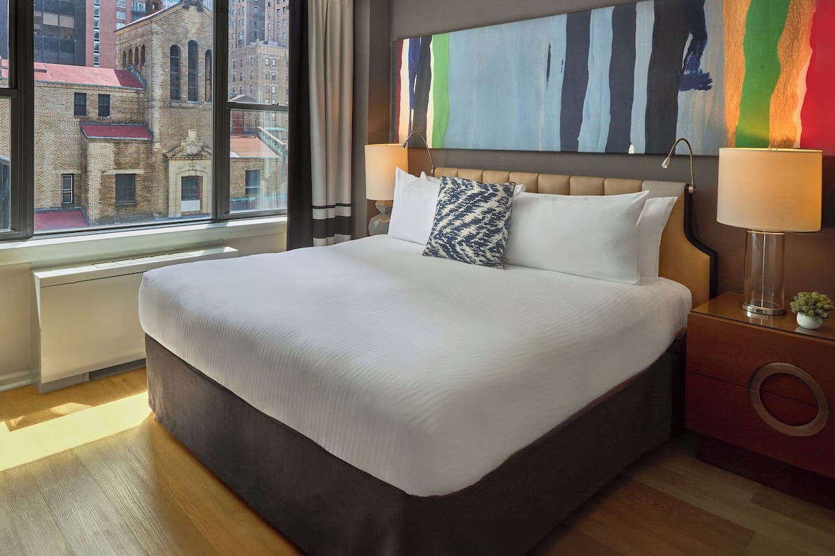 Quarto de hotel com uma cama grande, vista da cidade pela janela, pintura abstrata moderna na parede e mesa de cabeceira com abajur.