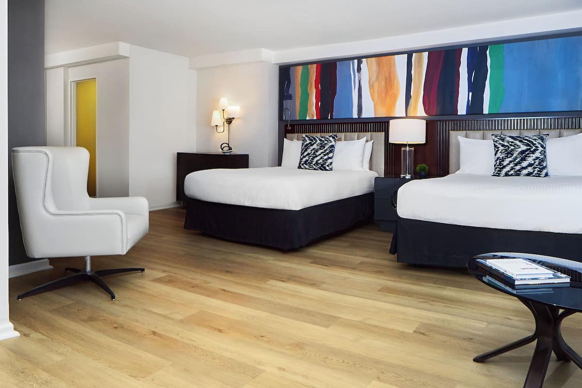 Quarto de hotel com duas camas, uma cadeira branca, uma mesinha pequena e um quadro abstrato colorido na parede.