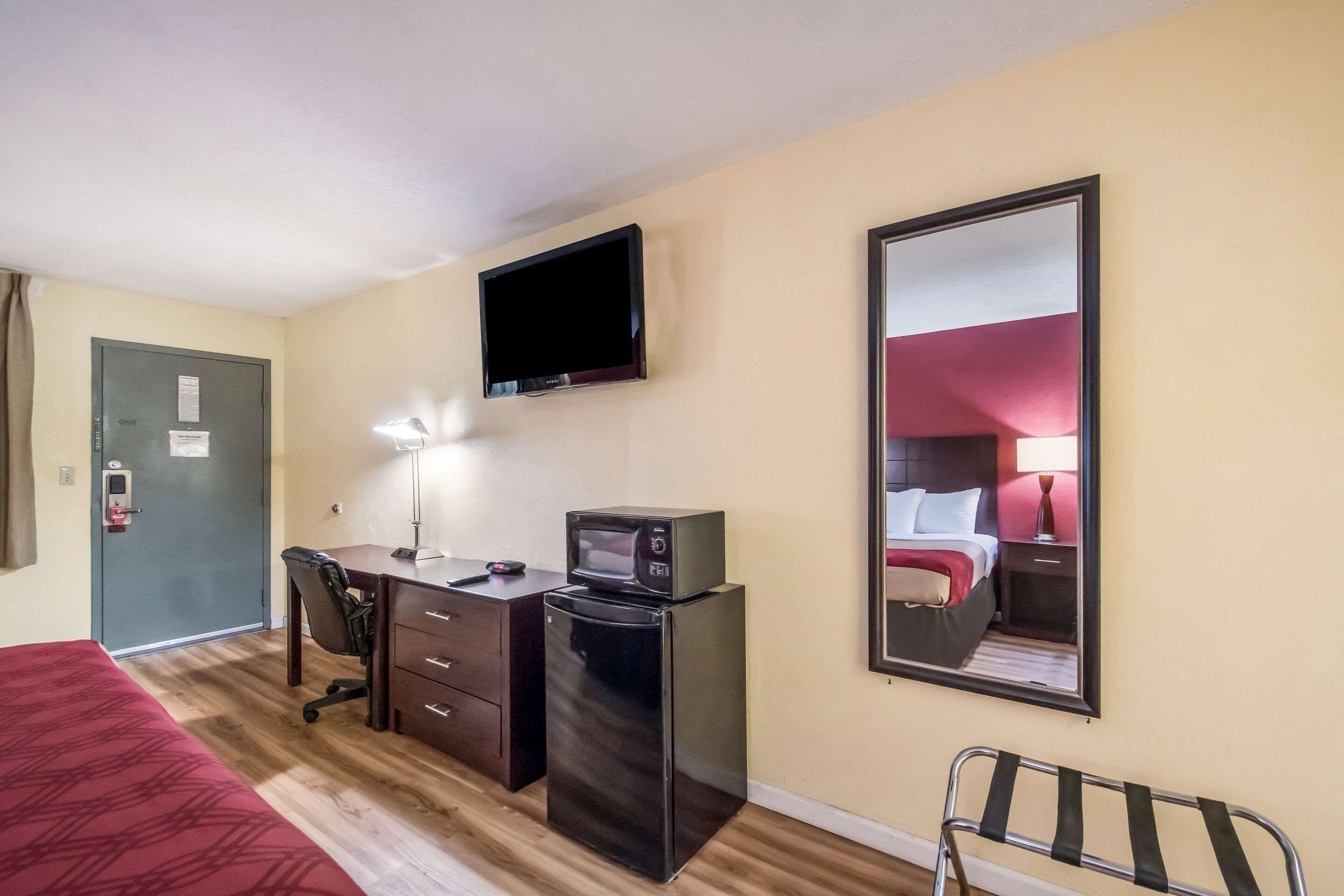Quarto de hotel com cama, escrivaninha com computador, micro-ondas, geladeira, televisão e espelho.