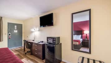 Quarto de hotel com cama, escrivaninha com computador, micro-ondas, geladeira, televisão e espelho.