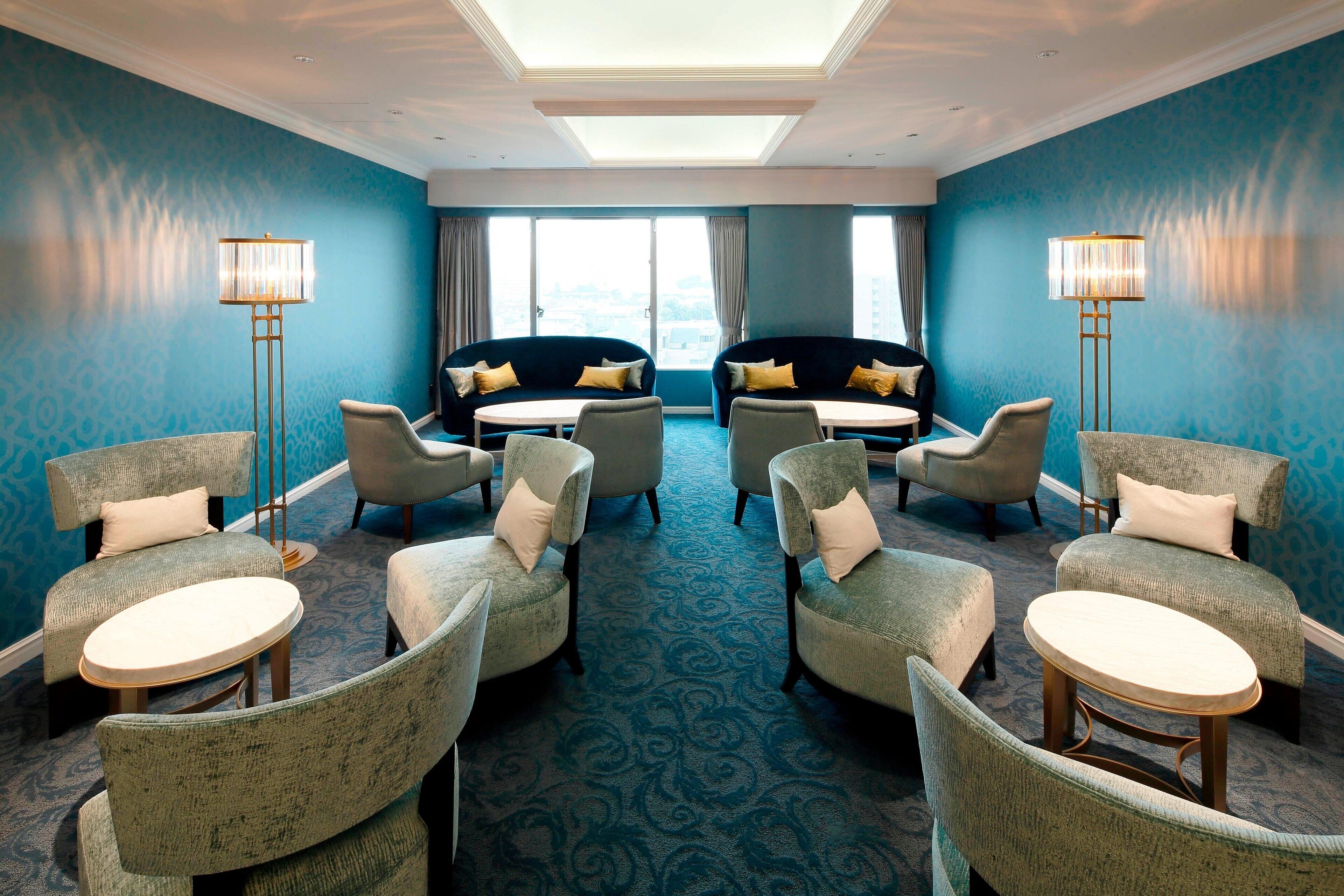 Quarto de hotel com carpete azul, dois sofás, quatro poltronas, duas mesinhas redondas e luminárias de chão.