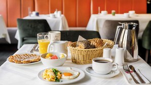 Uma mesa posta para o café da manhã, com waffles, ovos mexidos, uma tigela de frutas, uma cesta de pão, uma xícara de café e um copo de suco.