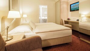 Quarto de hotel com uma cama, duas mesas de cabeceira, uma escrivaninha com cadeira, televisão fixada na parede e um banheiro visível através de uma porta aberta.