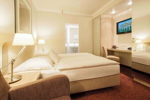 Quarto de hotel com uma cama, duas mesas de cabeceira, uma escrivaninha com cadeira, televisão fixada na parede e um banheiro visível através de uma porta aberta.