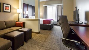 Quarto de hotel com sofá, escrivaninha com computador, cama, televisão e janela com cortinas.