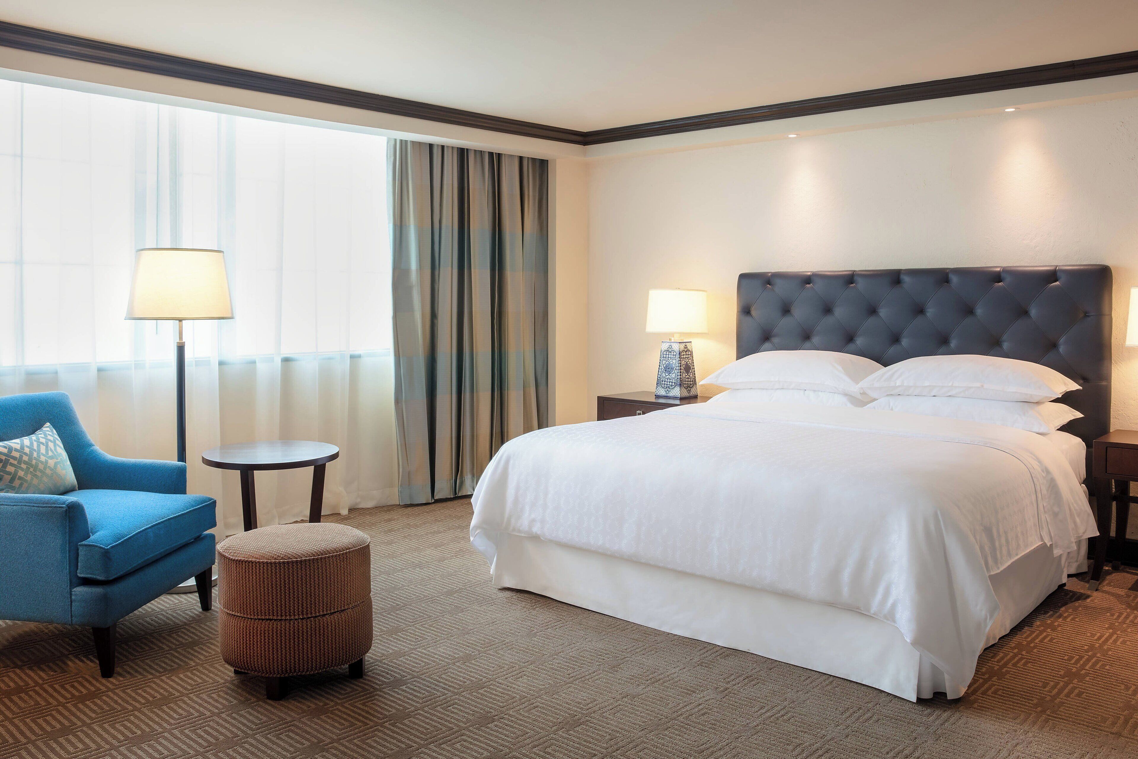 Quarto de hotel com uma cama grande, uma poltrona azul, uma mesinha redonda e um banquinho redondo.