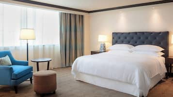 Quarto de hotel com uma cama grande, uma poltrona azul, uma mesinha redonda e um banquinho redondo.