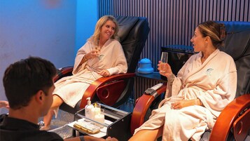 Duas pessoas em um quarto com cadeiras de massagem, uma mesinha com uma vela e um objeto azul em uma mesinha lateral.