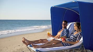 Um casal relaxa em uma cadeira de praia sob um toldo azul, com o oceano ao fundo.