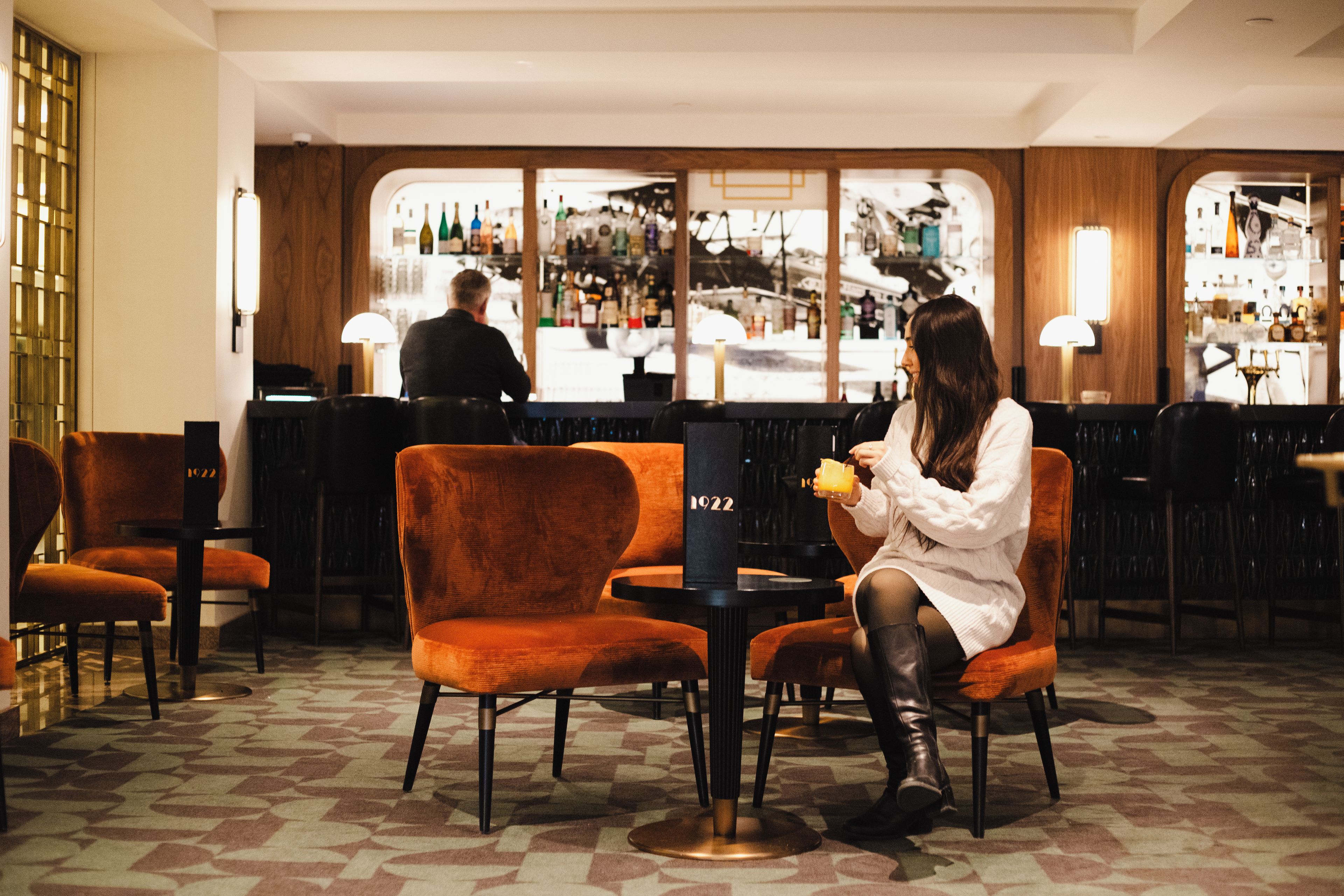 Uma área de bar com uma mulher sentada a uma mesa, um bartender ao fundo e um balcão de bar com vários frascos.