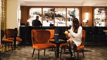 Uma área de bar com uma mulher sentada a uma mesa, um bartender ao fundo e um balcão de bar com vários frascos.