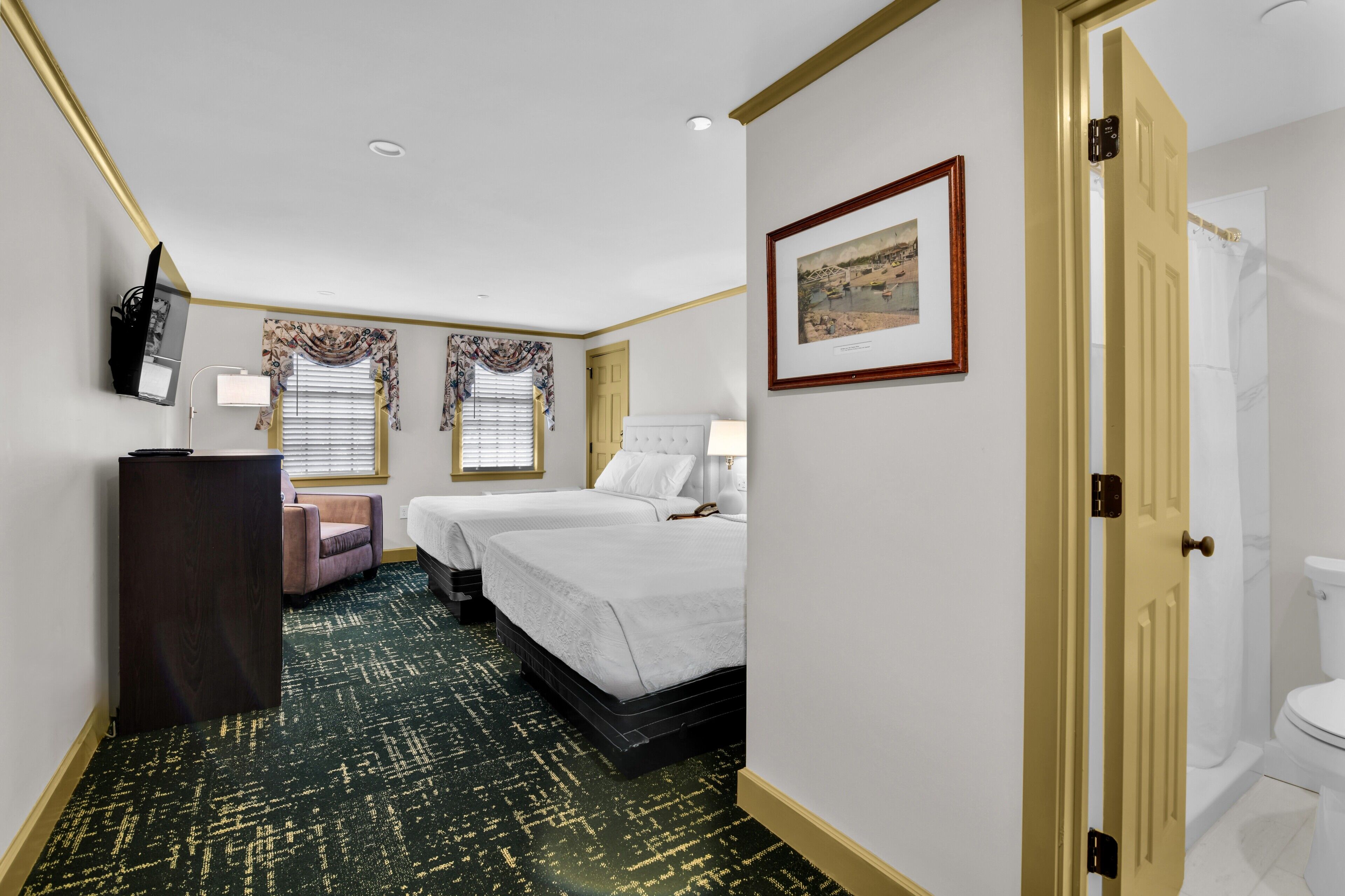 Quarto de hotel com duas camas, uma escrivaninha, uma cadeira, um quadro pendurado na parede e banheiro com vaso sanitário e chuveiro.