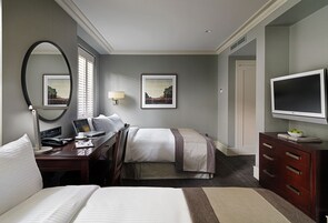 Quarto de hotel com duas camas, uma escrivaninha com computador, televisão e obras de arte nas paredes.
