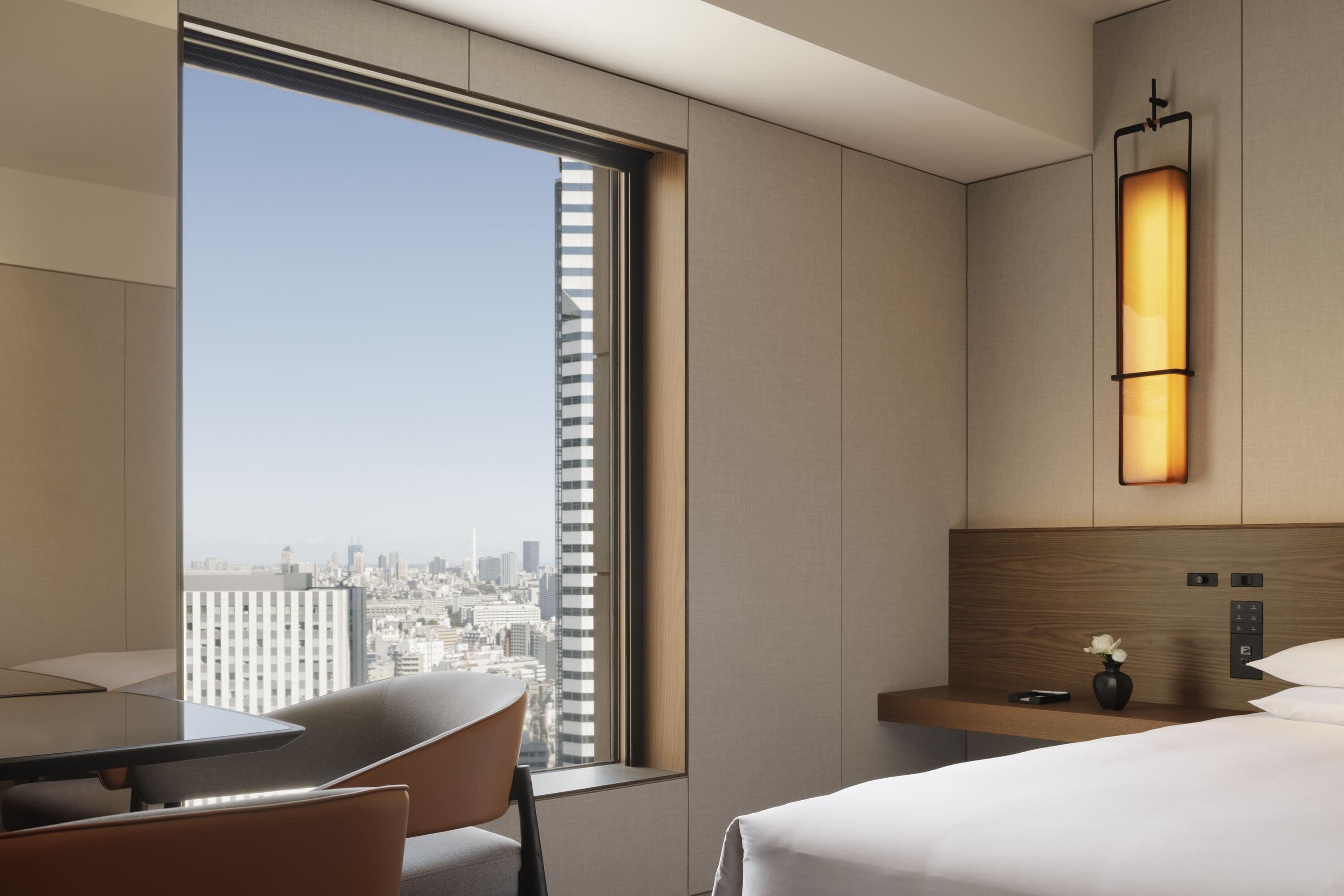 Quarto de hotel com uma janela grande, uma cama, uma mesa de cabeceira, uma cadeira e luminária fixada na parede.