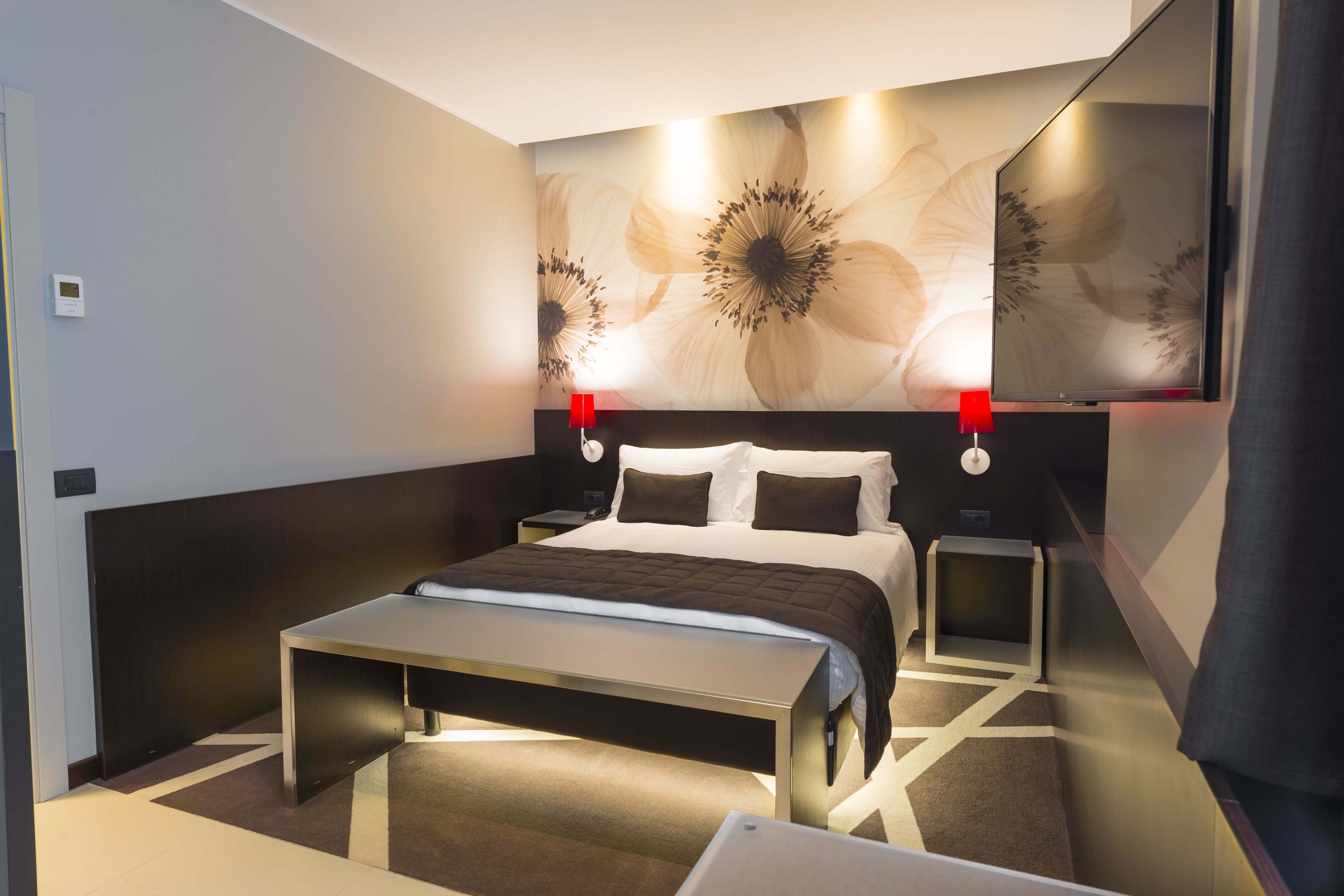 Quarto moderno com cama grande, mesas de cabeceira, um banquinho e televisão fixada na parede.