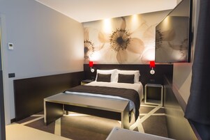 Quarto moderno com cama grande, mesas de cabeceira, um banquinho e televisão fixada na parede.