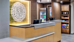 Uma área de recepção com balcão de madeira, um painel decorativo, um refrigerador com bebidas e um micro-ondas sobre uma prateleira com lanches.