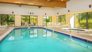Piscina com água azul cristalina, rodeada por espreguiçadeiras e guarda-sóis, com grandes janelas que oferecem vista para o verde.