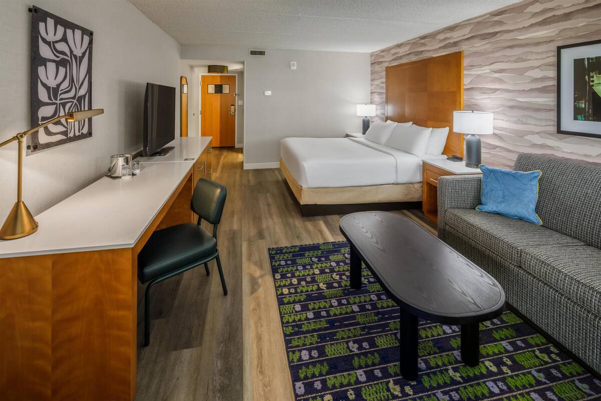 Quarto de hotel com cama, escrivaninha com cadeira, sofá, mesa de centro, televisão e luminária.