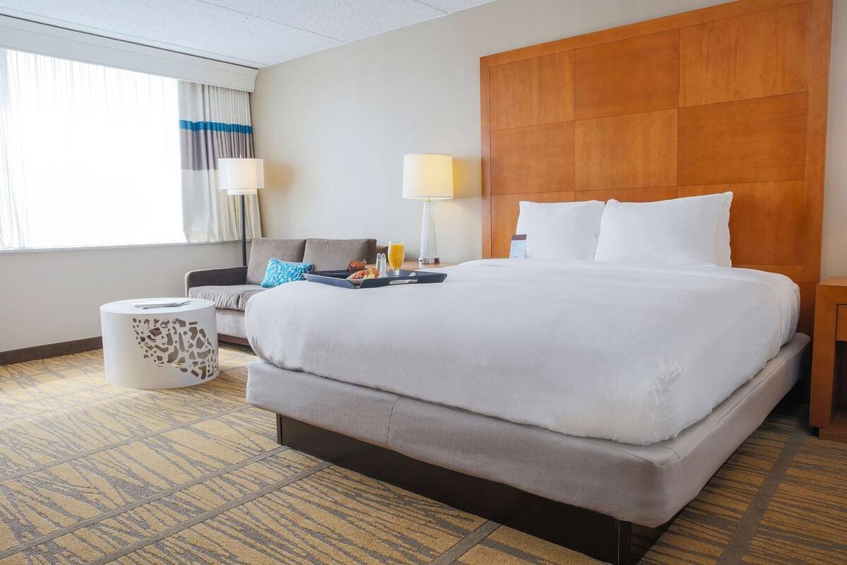 Quarto de hotel com uma cama grande, um sofá, uma mesa de apoio com uma luminária, uma pequena mesa redonda com um objeto decorativo e uma janela com cortinas.