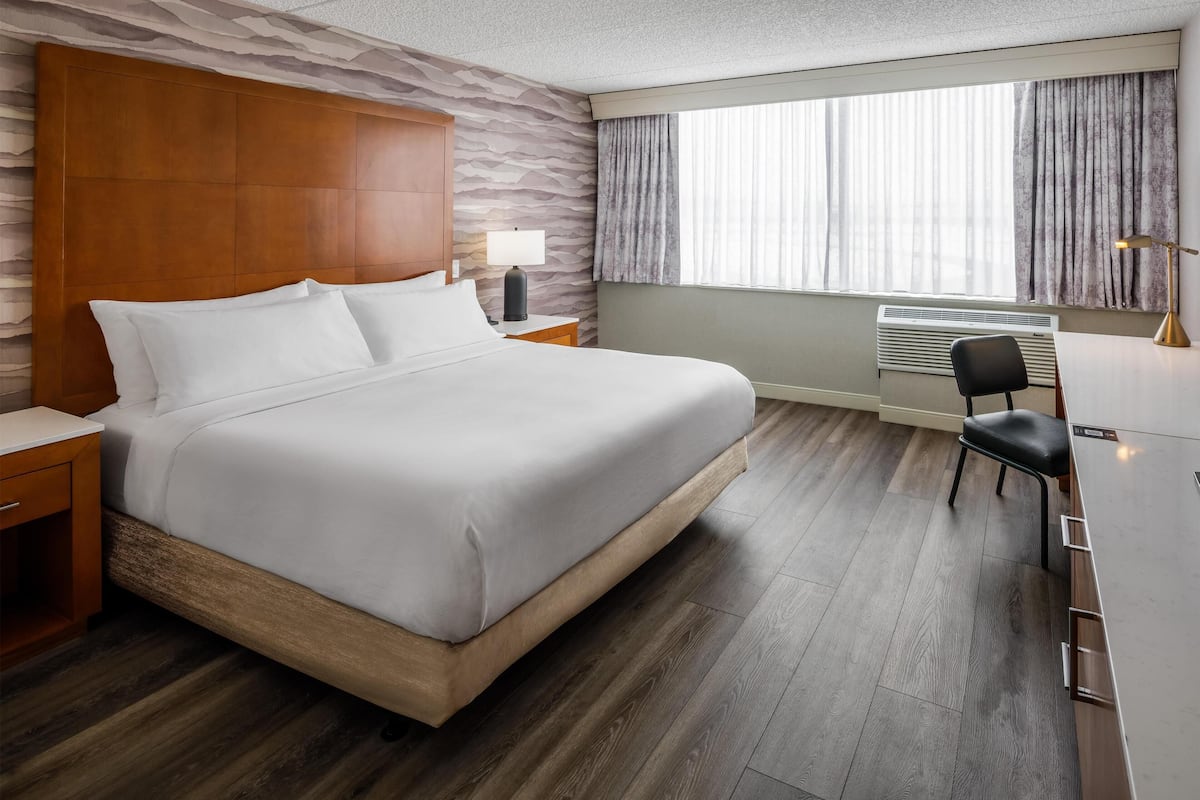 Quarto de hotel com cama de cabeceira em madeira, escrivaninha com cadeira, luminária e janela com cortinas.