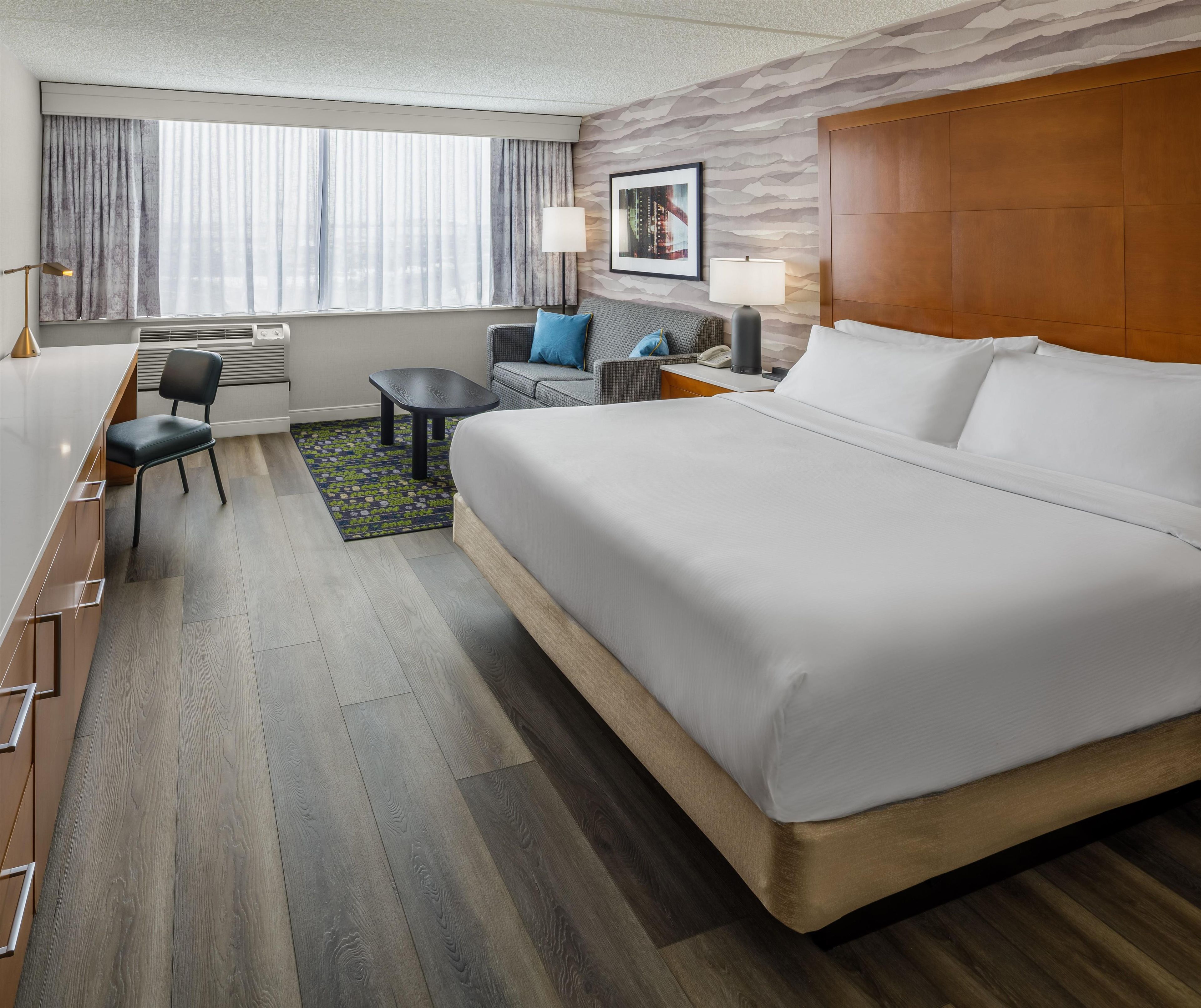 Quarto de hotel com uma cama grande, uma escrivaninha, uma cadeira, uma mesinha, um abajur e um quadro na parede.