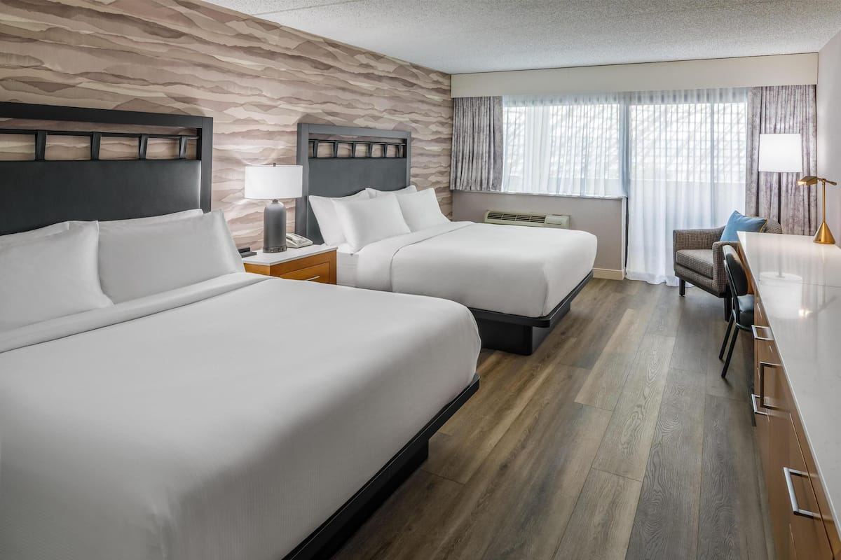 Quarto de hotel com duas camas, uma escrivaninha, uma cadeira, um abajur e uma janela com cortinas.