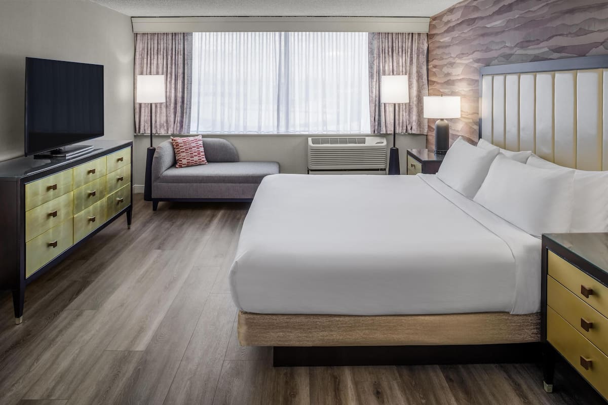 Quarto de hotel com uma cama grande, televisão, sofá, escrivaninha e janela com cortinas.