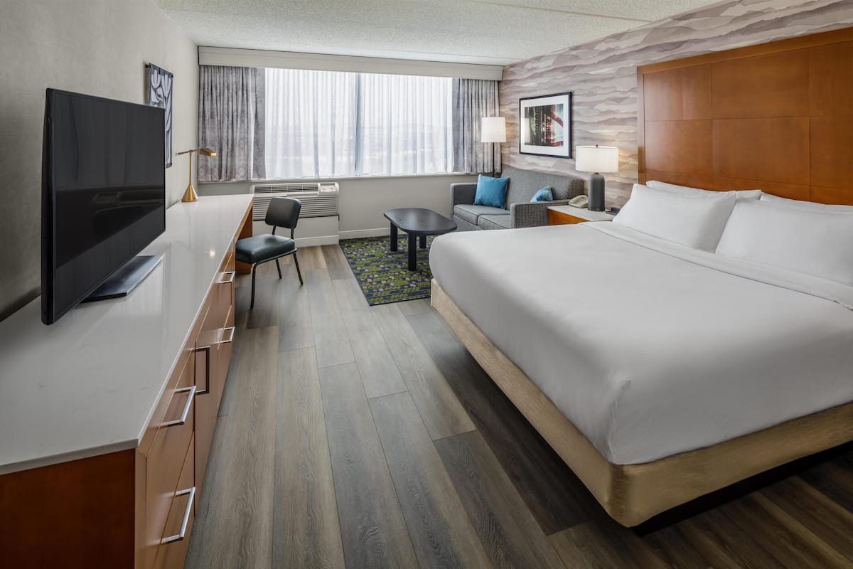 Quarto de hotel com cama, televisão, escrivaninha com cadeira, mesa pequena e sofá.