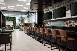 Uma área de bar moderna com mesas altas, banquetas e grandes janelas, além de um balcão de bar visível com diversos tipos de bebidas.