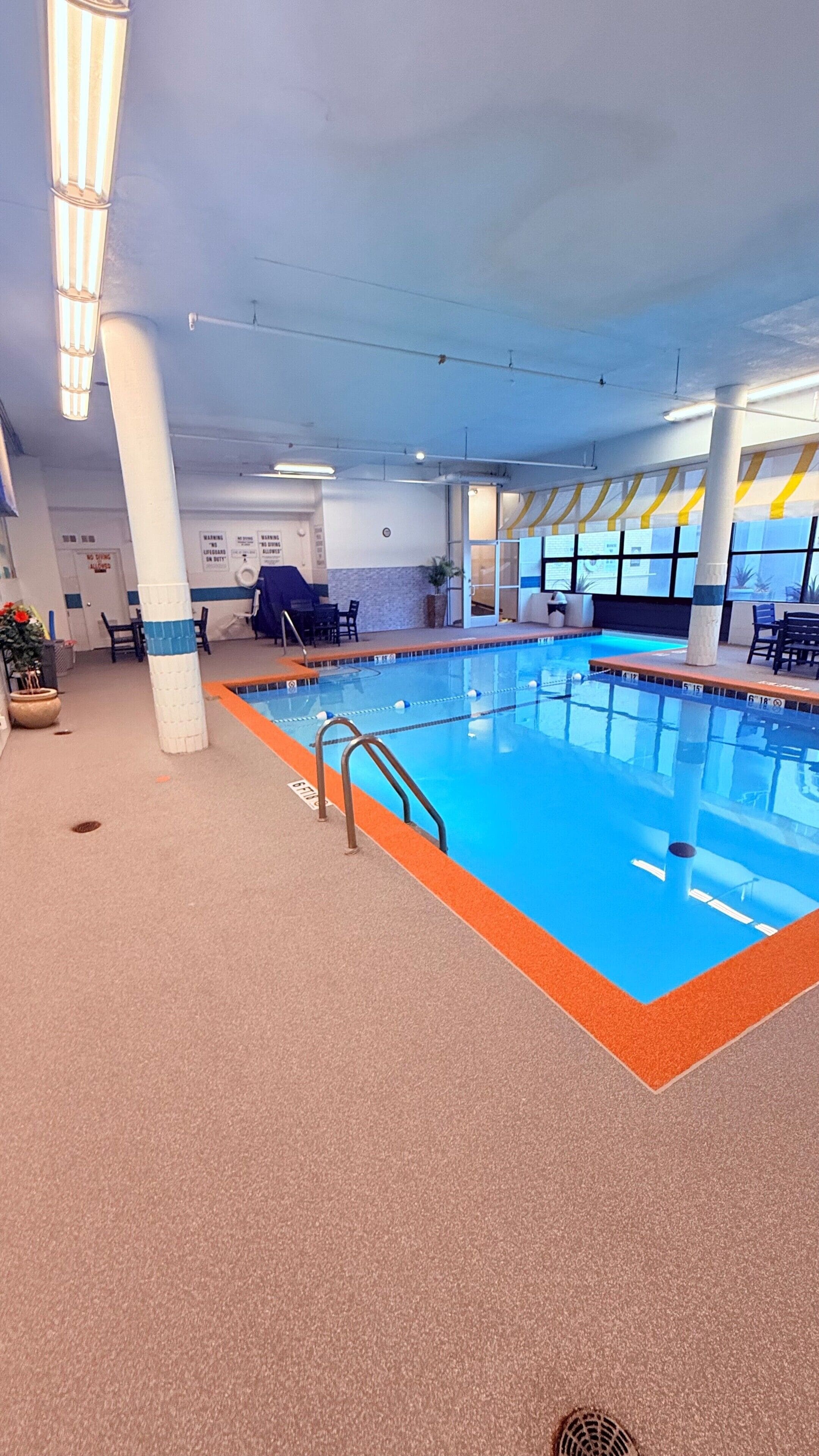Uma piscina interior com superfície de água cristalina, rodeada por um deck revestido em azulejos, e uma escada de acesso à piscina.