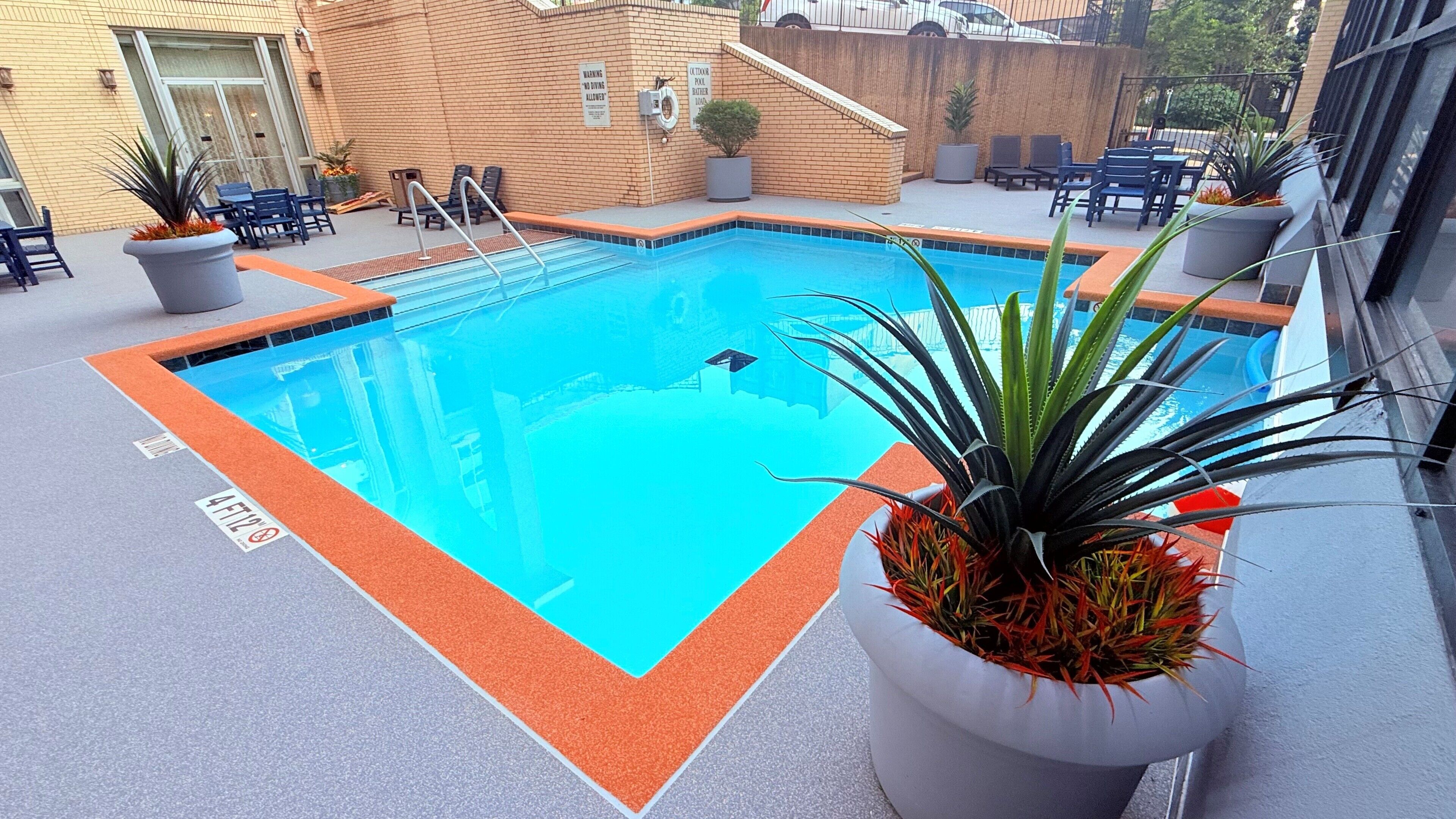 Uma piscina retangular com interior azul, rodeada por uma borda laranja texturizada. Há espreguiçadeiras e plantas em vasos ao redor da piscina.