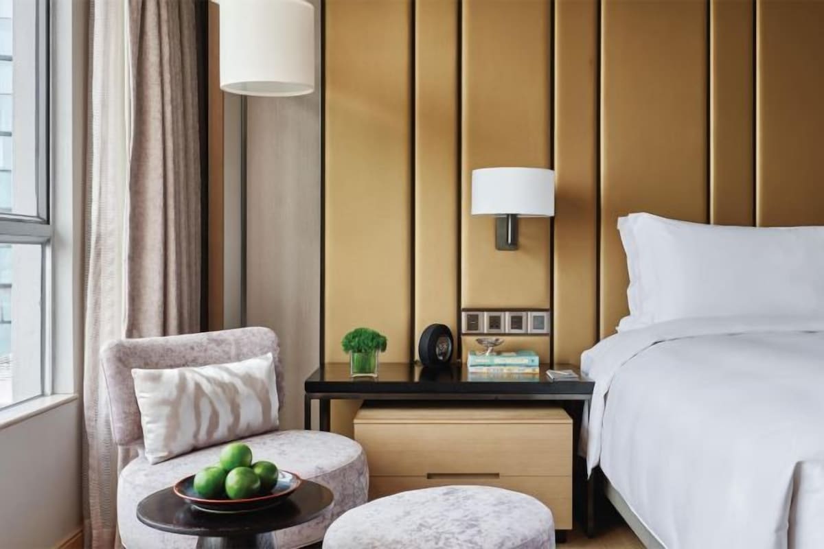 Quarto de hotel com cama, mesa de cabeceira, abajur, cadeira, pequena mesa com uma tigela de frutas e janela com cortinas.