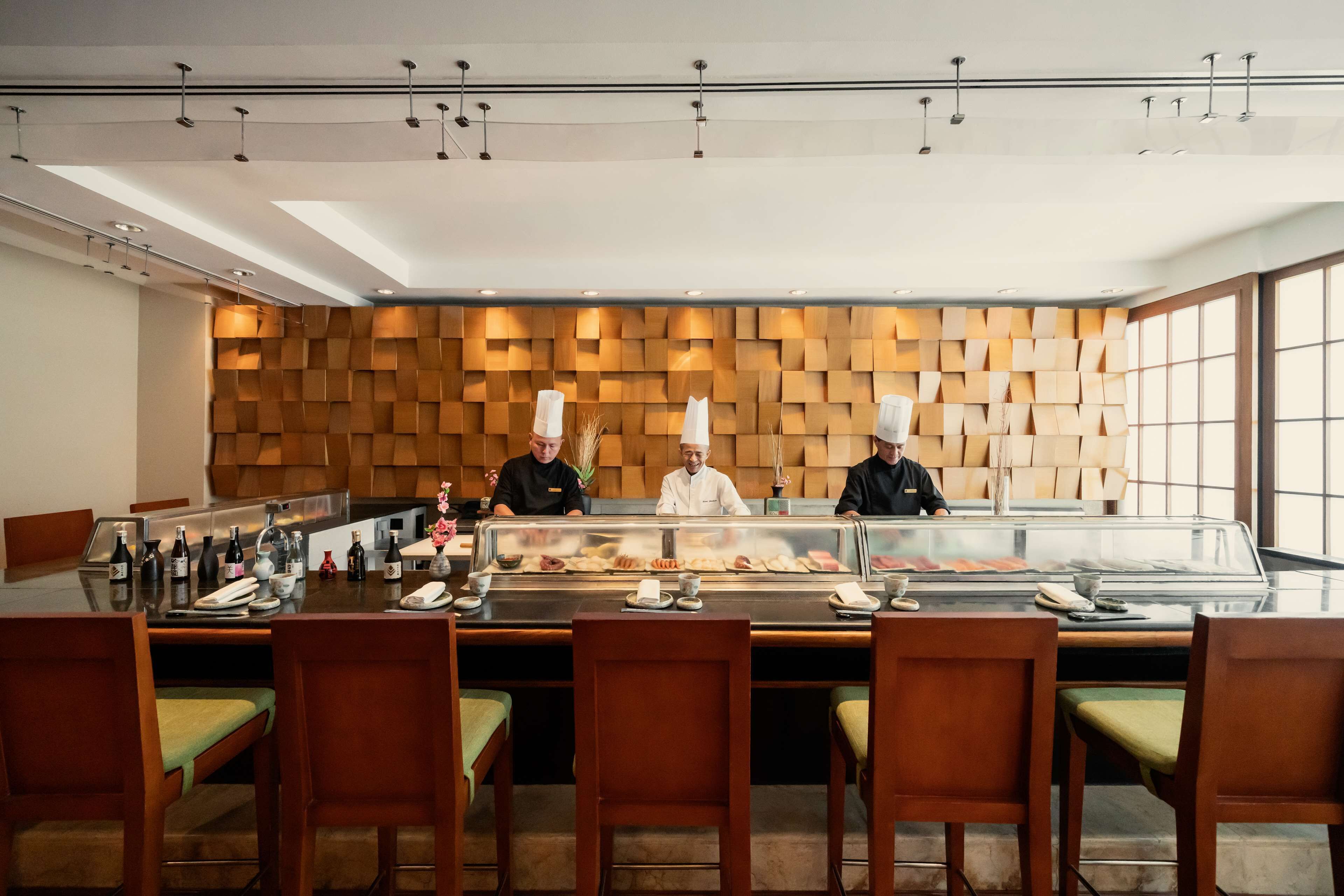 Um restaurante moderno com um balcão central, três funcionários e uma variedade de pratos expostos numa vitrine.
