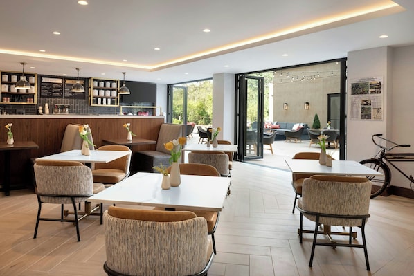 Uma área de refeições moderna com mesas e cadeiras, um espaço de bar com prateleiras e vista para o exterior.