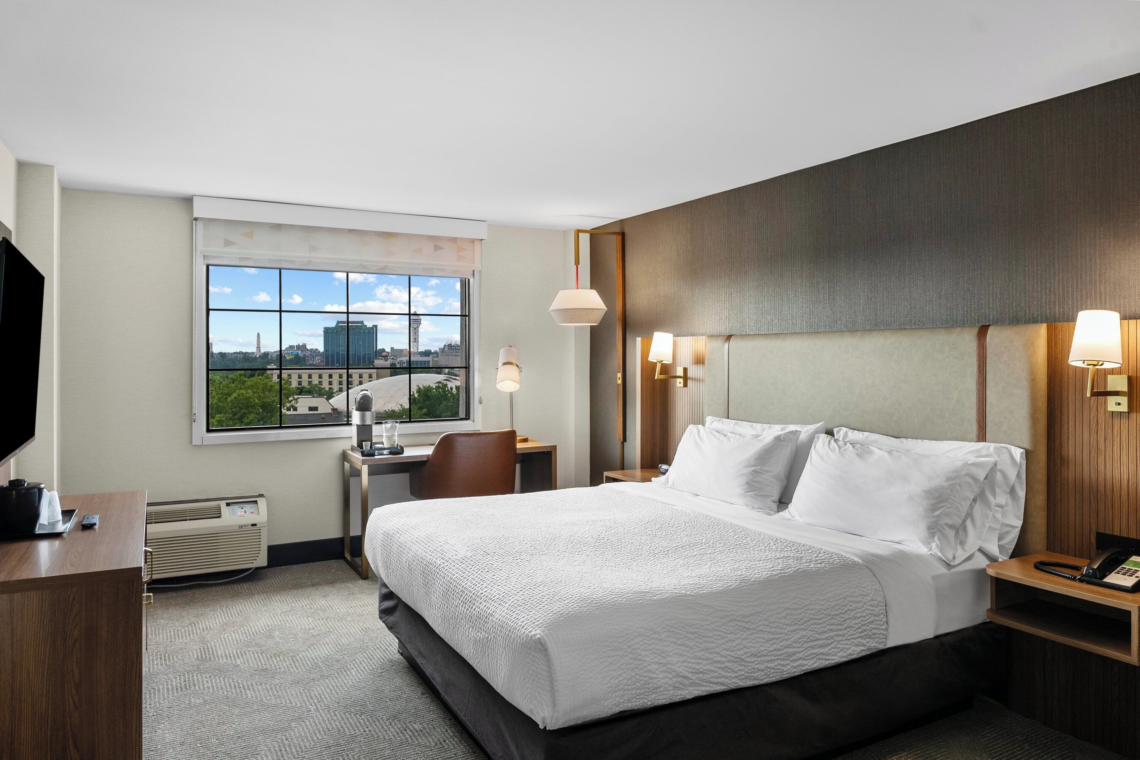 Quarto de hotel com uma cama grande, uma escrivaninha, uma cadeira, uma televisão, uma janela com vista para a cidade e uma luminária de parede.