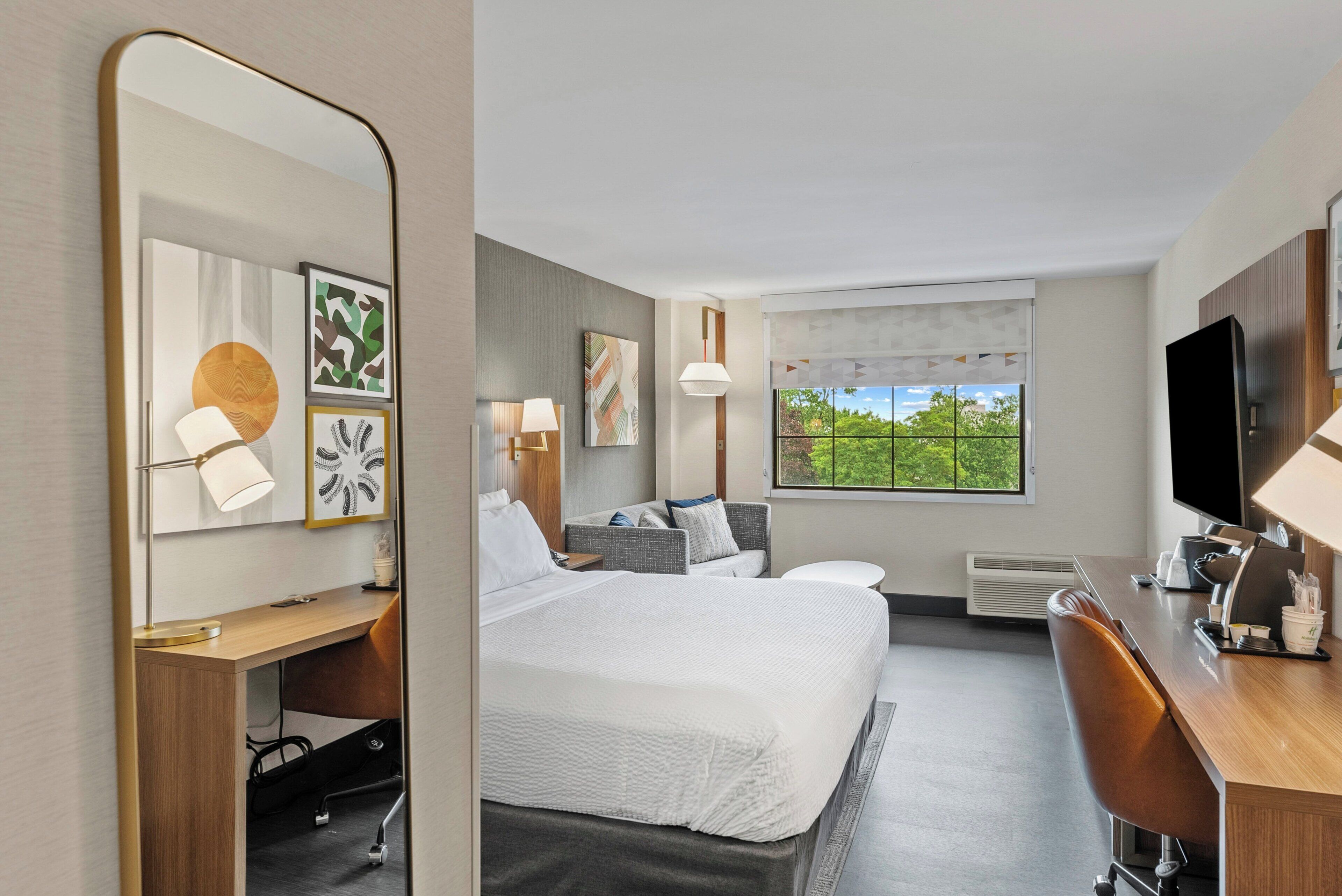 Quarto de hotel moderno com uma cama grande, uma escrivaninha, uma cadeira, televisão e janela com vista para o verde.