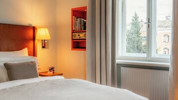 Quarto de hotel com cama, luminária de cabeceira, estante de livros e janela com vista para prédios e árvores.
