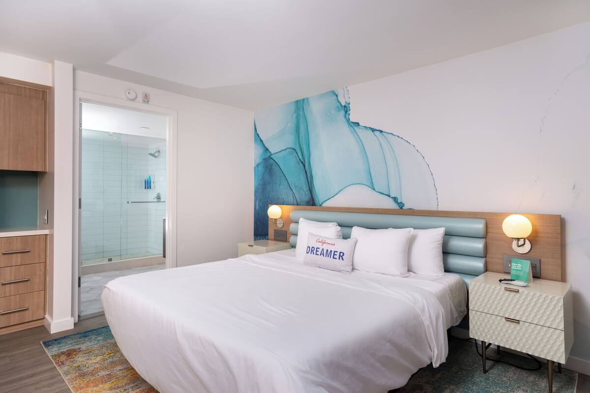Quarto de hotel com uma cama grande, uma mesa de cabeceira com abajur, uma obra de arte na parede e um banheiro visível através de uma porta aberta.