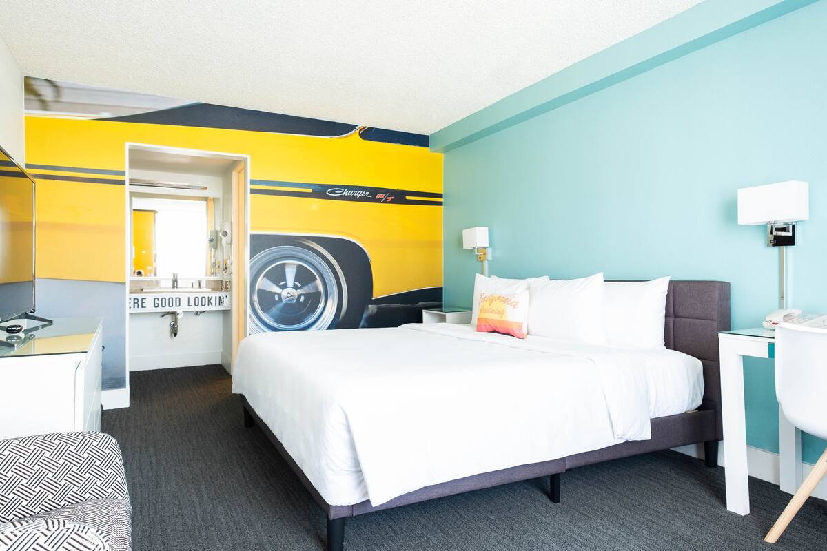 Quarto de hotel com cama, escrivaninha, cadeira, espelho e parede com adesivo de carro.